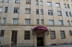 Fachada de hotel com toldo bordô, várias janelas e entrada com portão.