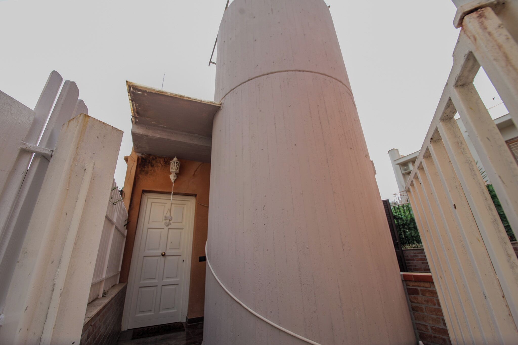 Uma entrada estreita com uma porta branca, cerca branca e uma estrutura cilíndrica grande.