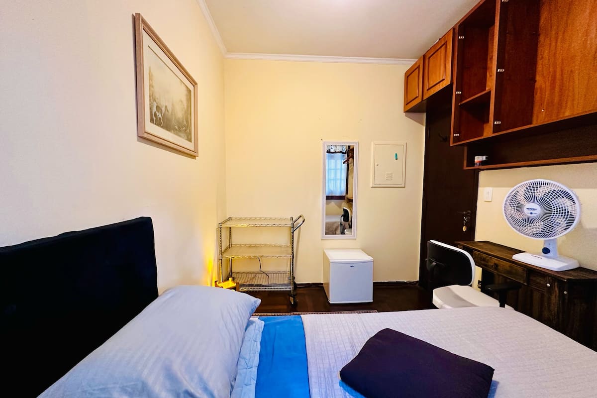 Quarto de hotel com cama, escrivaninha, cadeira, espelho, guarda-roupa e uma pequena cozinha.