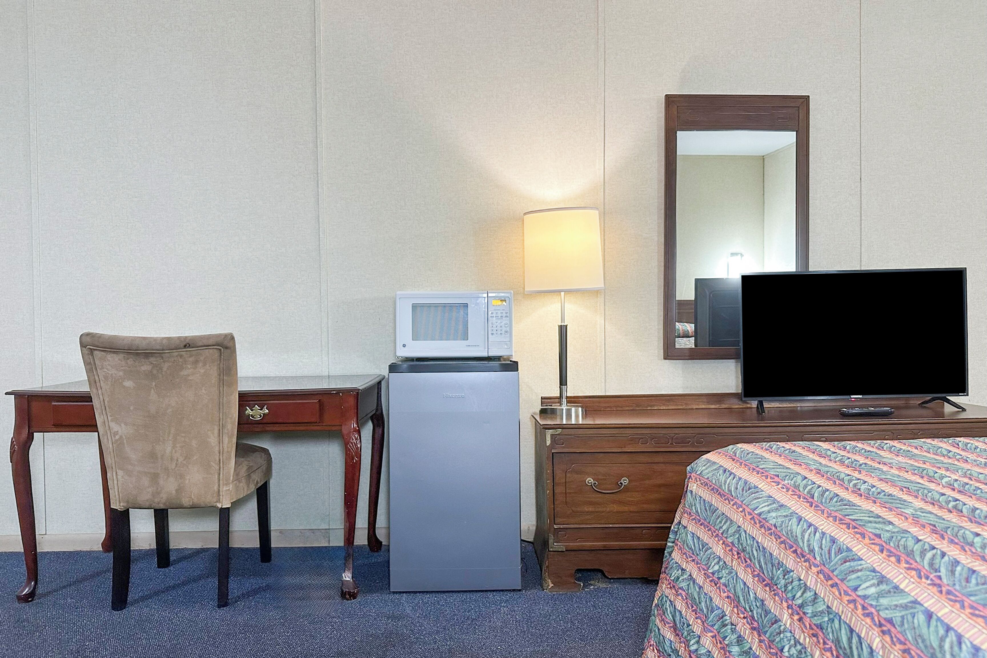 Quarto de hotel com cama, escrivaninha com micro-ondas, televisão, cadeira e cômoda.