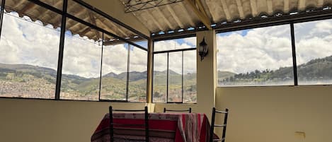 Um quarto com mesa e cadeiras, amplas janelas com vista para uma cidade e montanhas, e piso de azulejos.