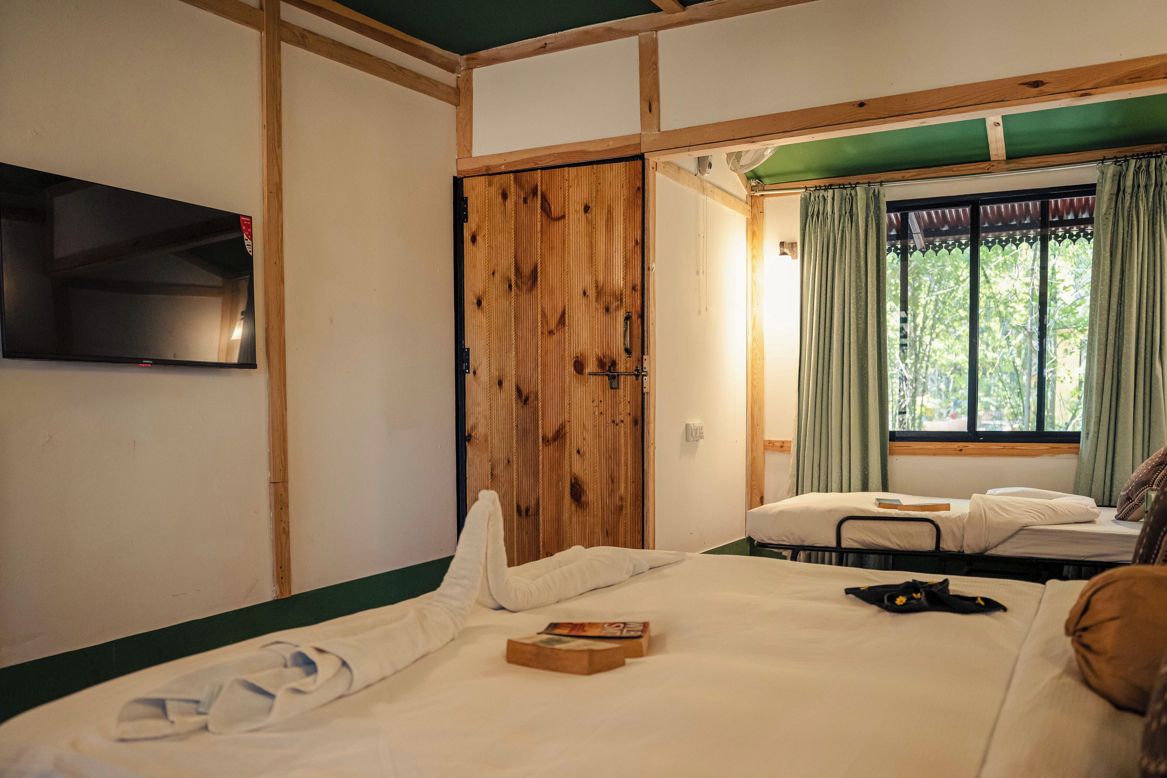 Quarto de hotel com guarda-roupa de madeira, cama com edredom branco, mesa de cabeceira com um livro e janela com cortinas verdes.