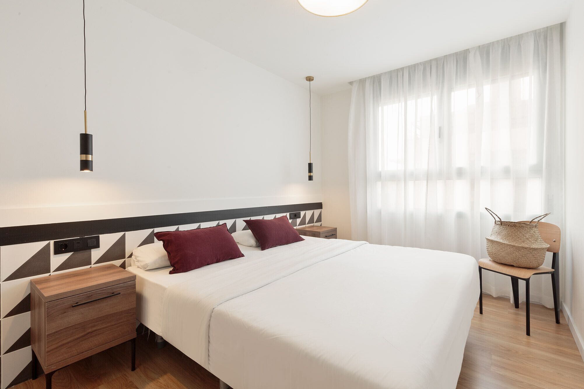 Quarto de hotel moderno com uma cama grande, mesas de cabeceira, uma mesinha de cabeceira de madeira, uma cesta de palha numa cadeira e cortinas translúcidas.