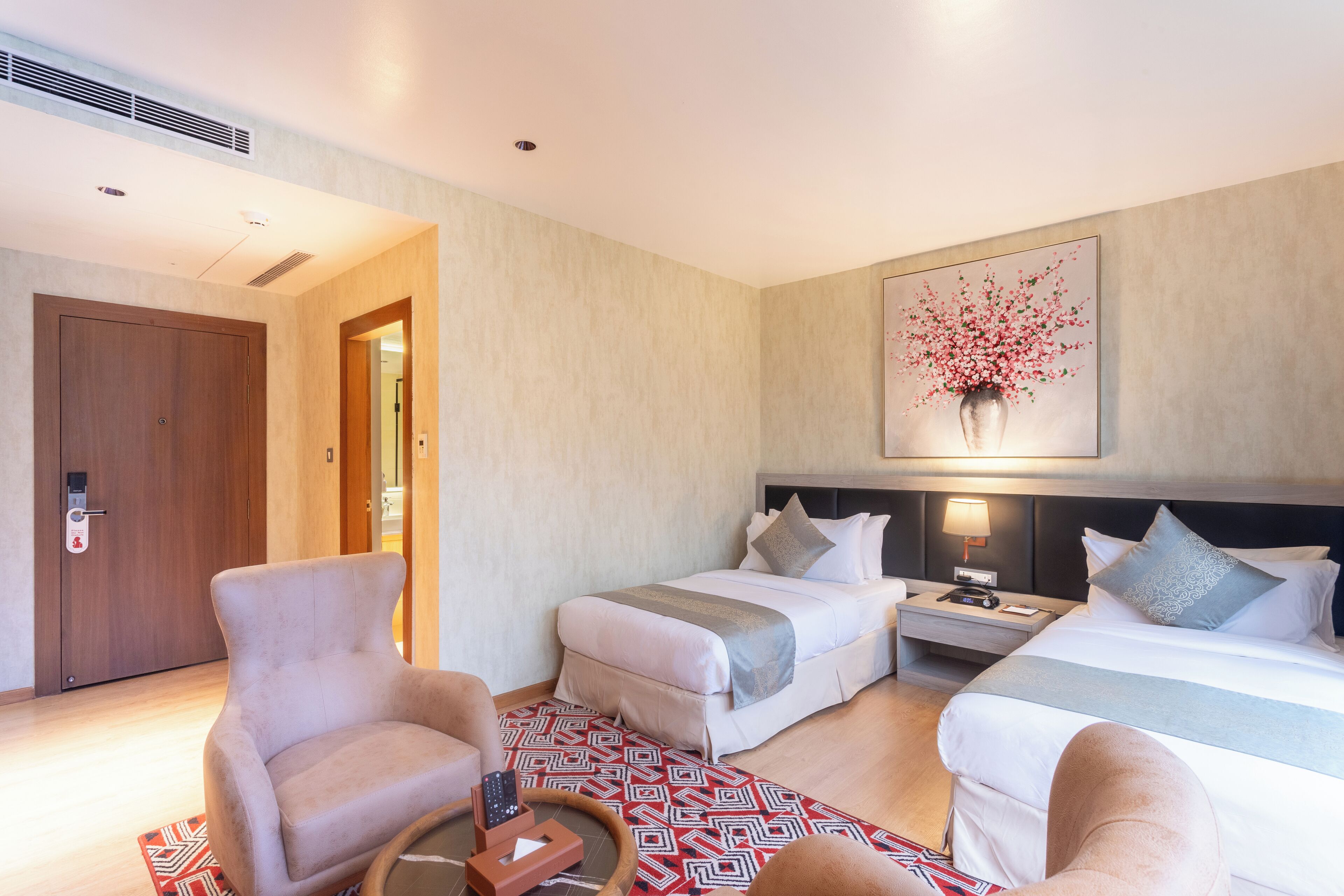 Quarto de hotel com duas camas, área de estar com uma cadeira, uma pequena mesa e um objeto decorativo na parede.