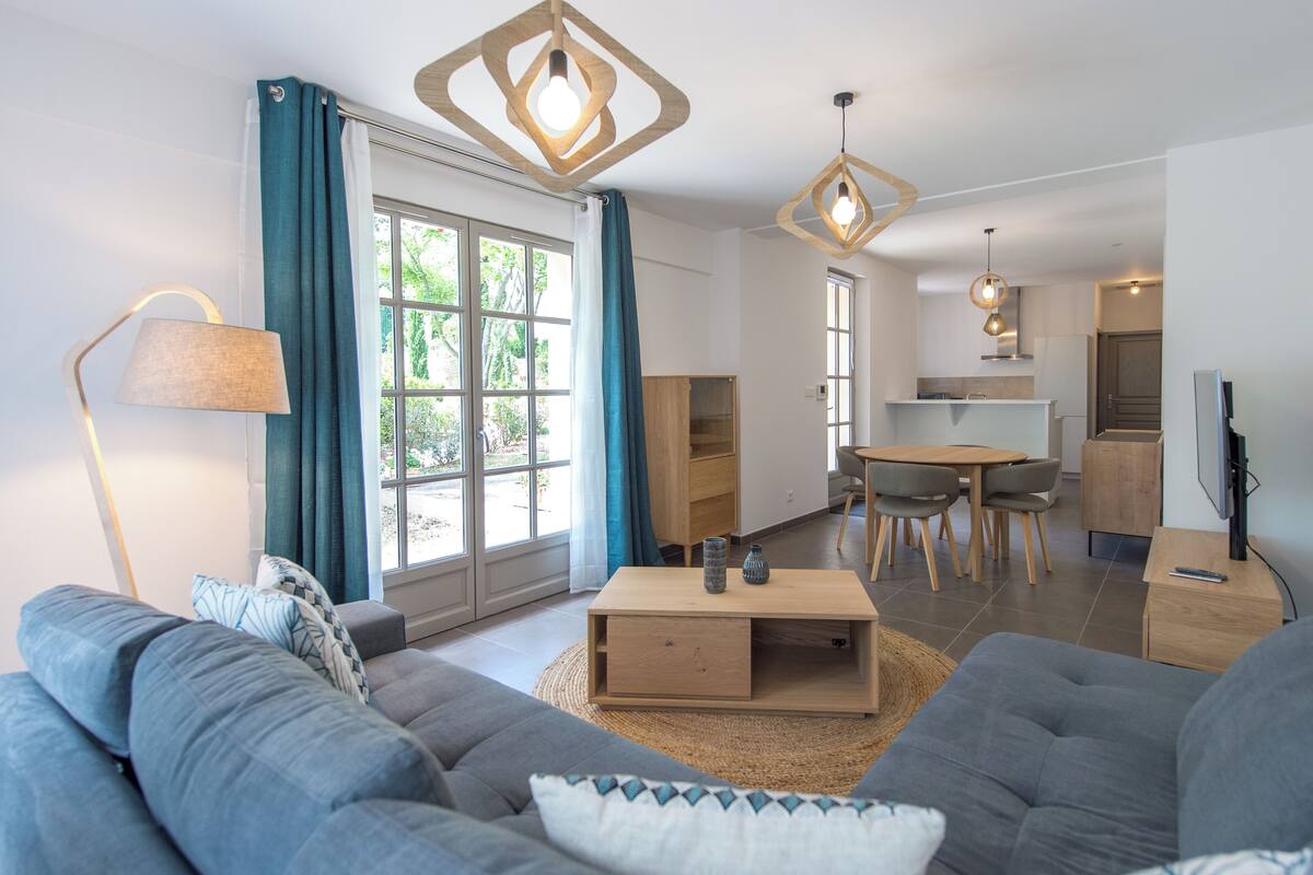 Uma sala de estar moderna com sofá, mesa de centro e luminárias pendentes. Há uma área de jantar com mesa e cadeiras, e uma cozinha ao fundo.