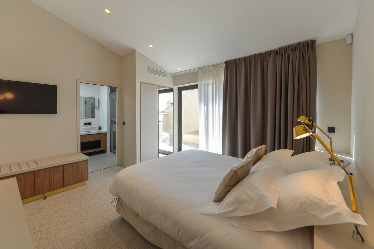 Quarto de hotel moderno com uma cama grande, televisão de tela plana, escrivaninha com cadeira e porta de correr para uma varanda.