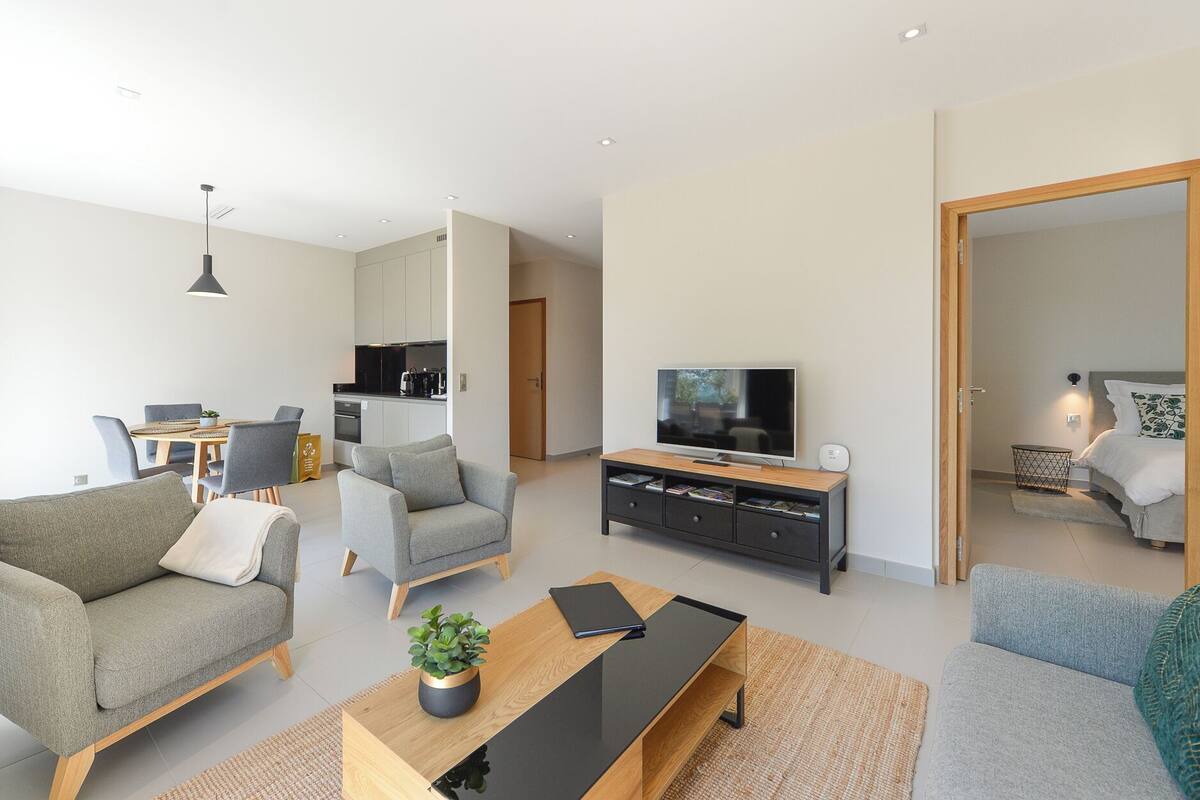 Uma sala de estar moderna com sofás cinzas, uma mesa de centro de madeira, uma televisão e uma cozinha ao fundo.