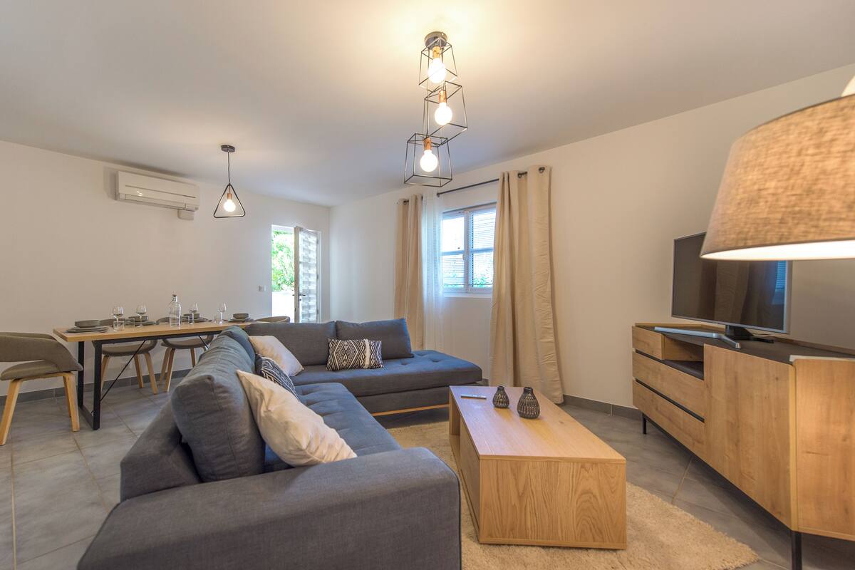Uma sala de estar moderna com um sofá seccional, uma mesa de centro de madeira, uma TV sobre um suporte de madeira e iluminação pendente.