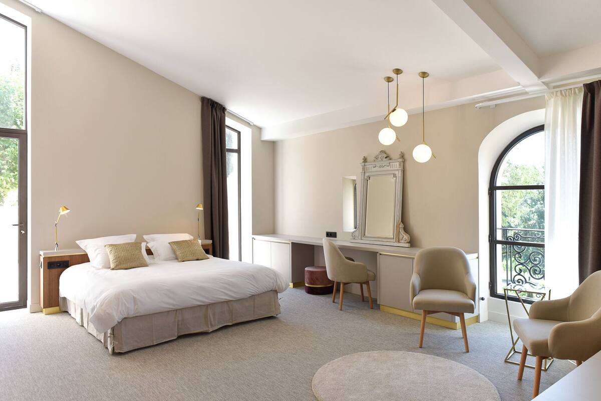 Um quarto espaçoso com uma cama grande, uma escrivaninha e uma cadeira, com um espelho amplo e vista para uma varanda.
