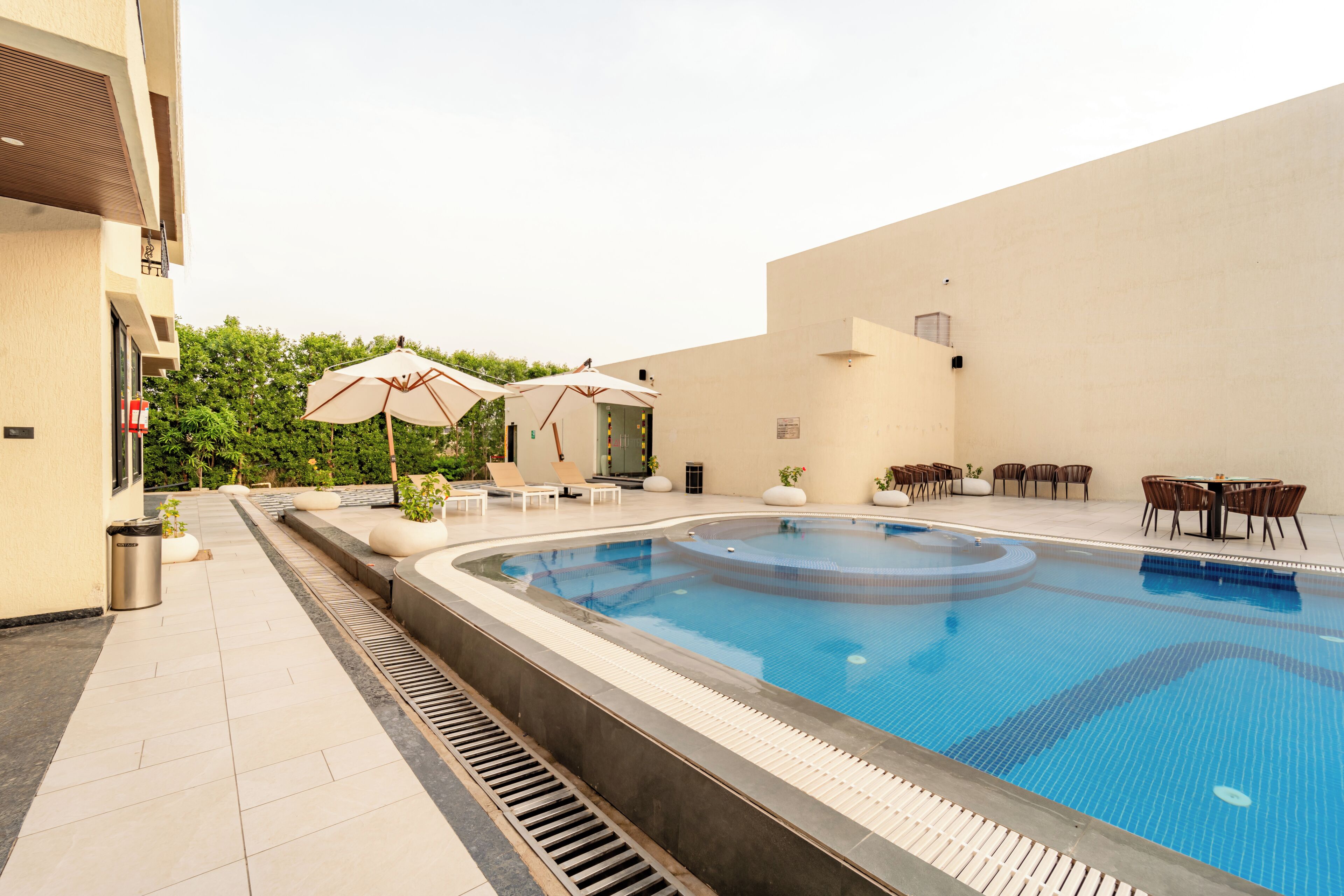 Uma piscina com espreguiçadeiras e guarda-sóis, rodeada por um edifício com chão de azulejos e uma lixeira.