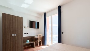 Quarto de hotel com cama, escrivaninha, cadeira, televisão, guarda-roupa e varanda com cortinas.