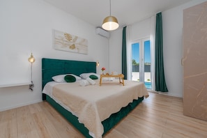 Quarto com cama estofada verde, mesa de cabeceira com bandeja, ar-condicionado fixado na parede e janela com cortinas.