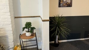 Um canto com parede de tijolos brancos, uma pequena mesa redonda com uma árvore bonsai, uma planta em vaso e uma pintura de flores.