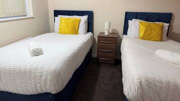 Quarto de hotel com duas camas, cada uma com cabeceira azul e roupa de cama branca. Há uma pequena mesa de cabeceira de madeira com uma luminária entre as camas.