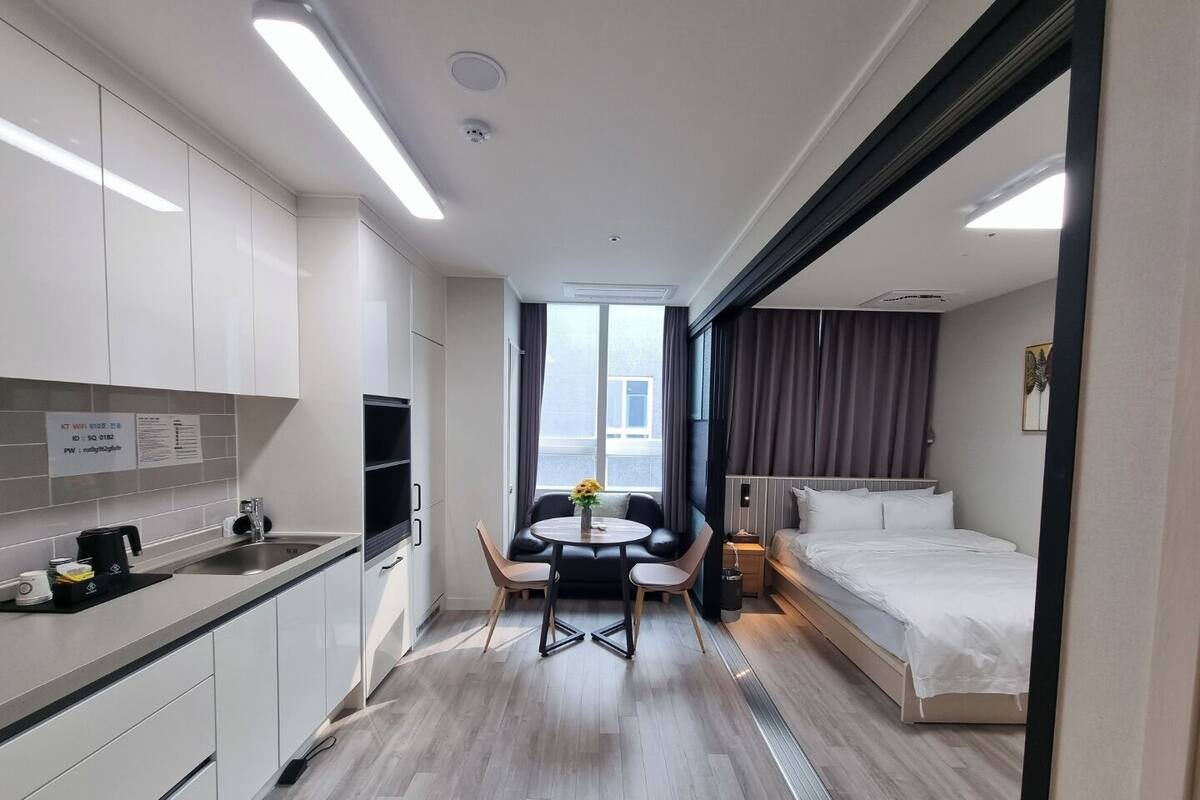 Quarto de hotel moderno com cozinha compacta, área de refeições com mesa e cadeiras, e uma cama grande com cabeceira.