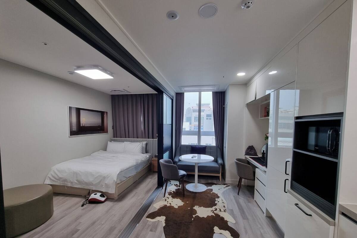 Quarto de hotel moderno com cama, escrivaninha, poltrona, sofá, televisão e uma grande janela.