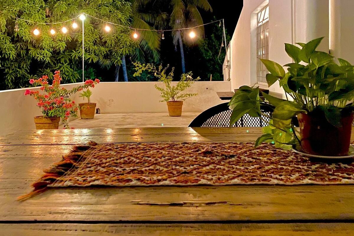 Área de estar ao ar livre com luzes de fada, plantas em vasos e mesa de madeira.