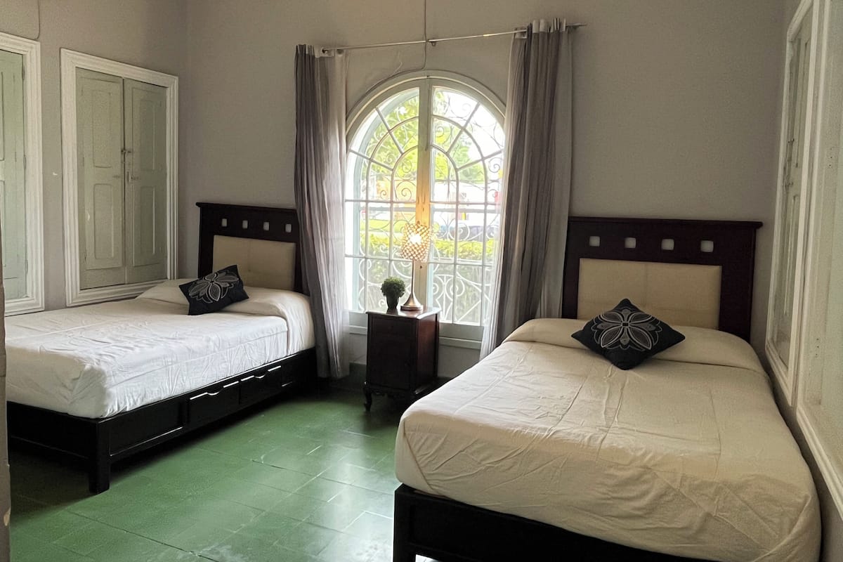 Quarto com duas camas, ventilador de teto, janela com cortinas e vista para o verde lá fora.