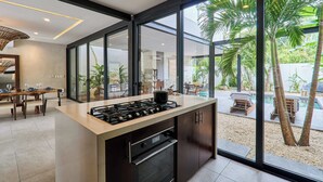 Uma cozinha moderna com uma ilha grande, uma área de refeições com mesa e cadeiras, e vista para uma piscina e palmeiras através de grandes janelas.