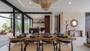 Uma sala de jantar moderna com uma longa mesa de madeira posta para seis pessoas, rodeada por cadeiras de vime.