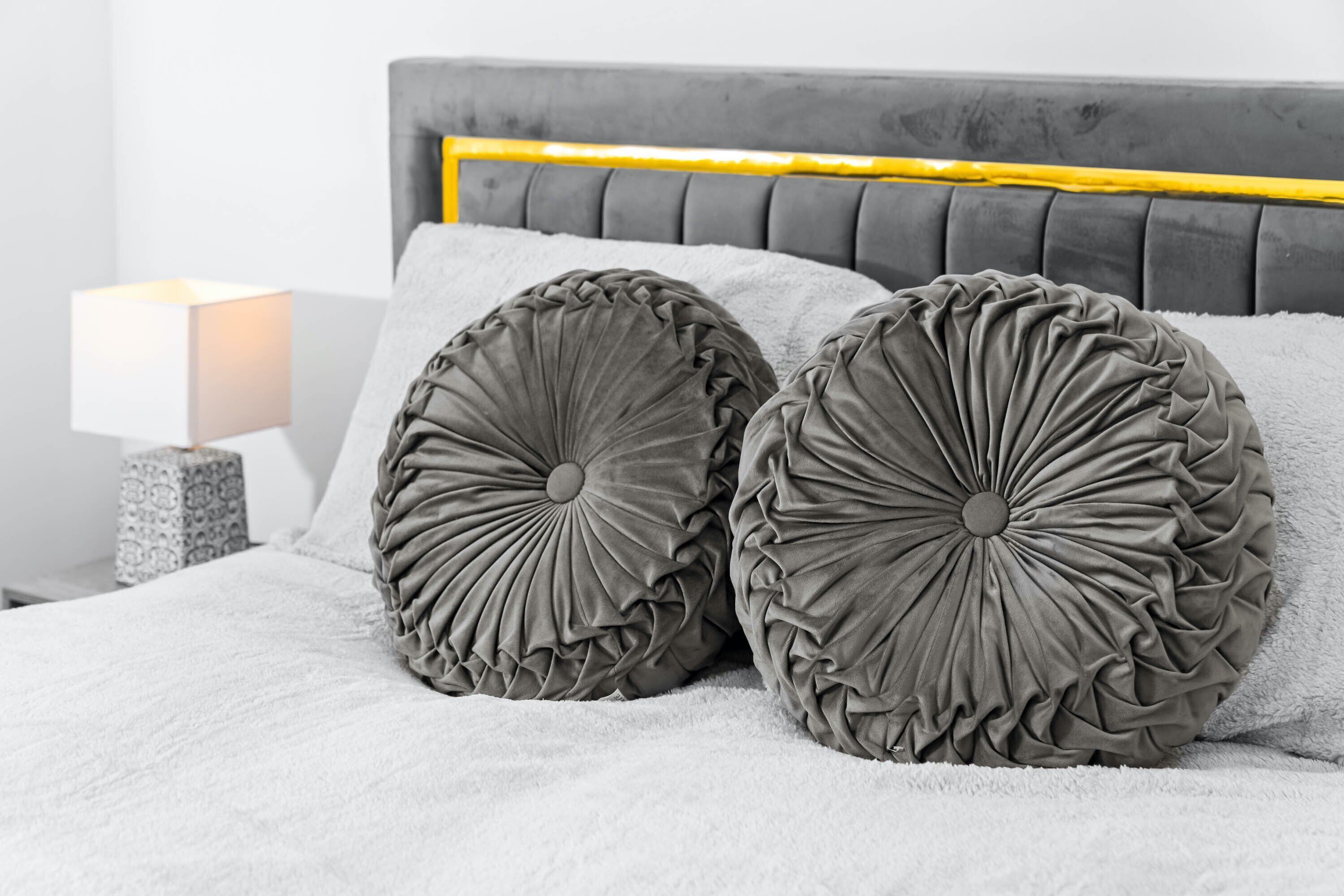 Uma cama com cabeceira estofada cinza, dois travesseiros cinzas com detalhes em babados, e uma mesa de cabeceira com uma luminária e um objeto decorativo.