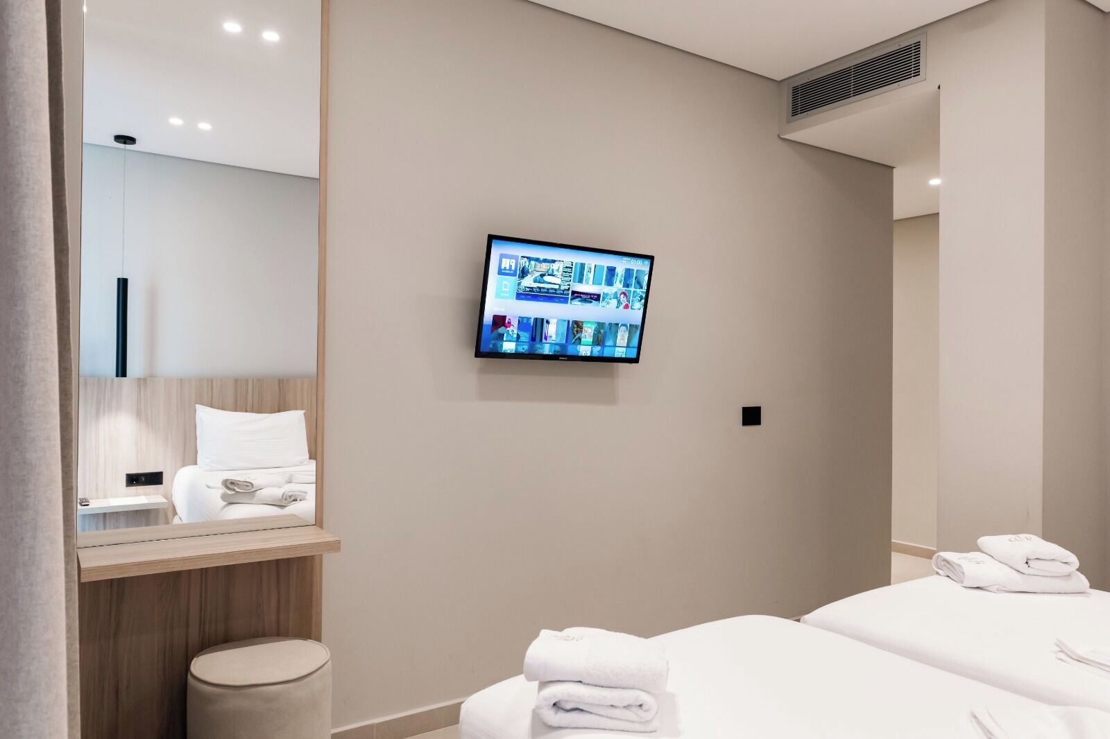 Quarto de hotel moderno com TV de tela plana na parede, penteadeira de madeira com espelho, um banquinho redondo e camas bem arrumadas com toalhas.