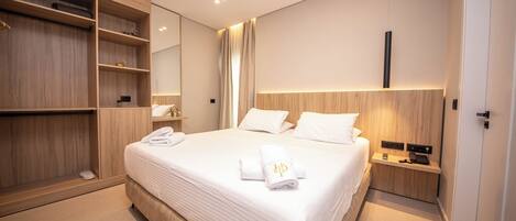 Quarto de hotel moderno com uma cama grande, um guarda-roupa de madeira, uma mesinha de cabeceira com abajur e um espelho na parede.