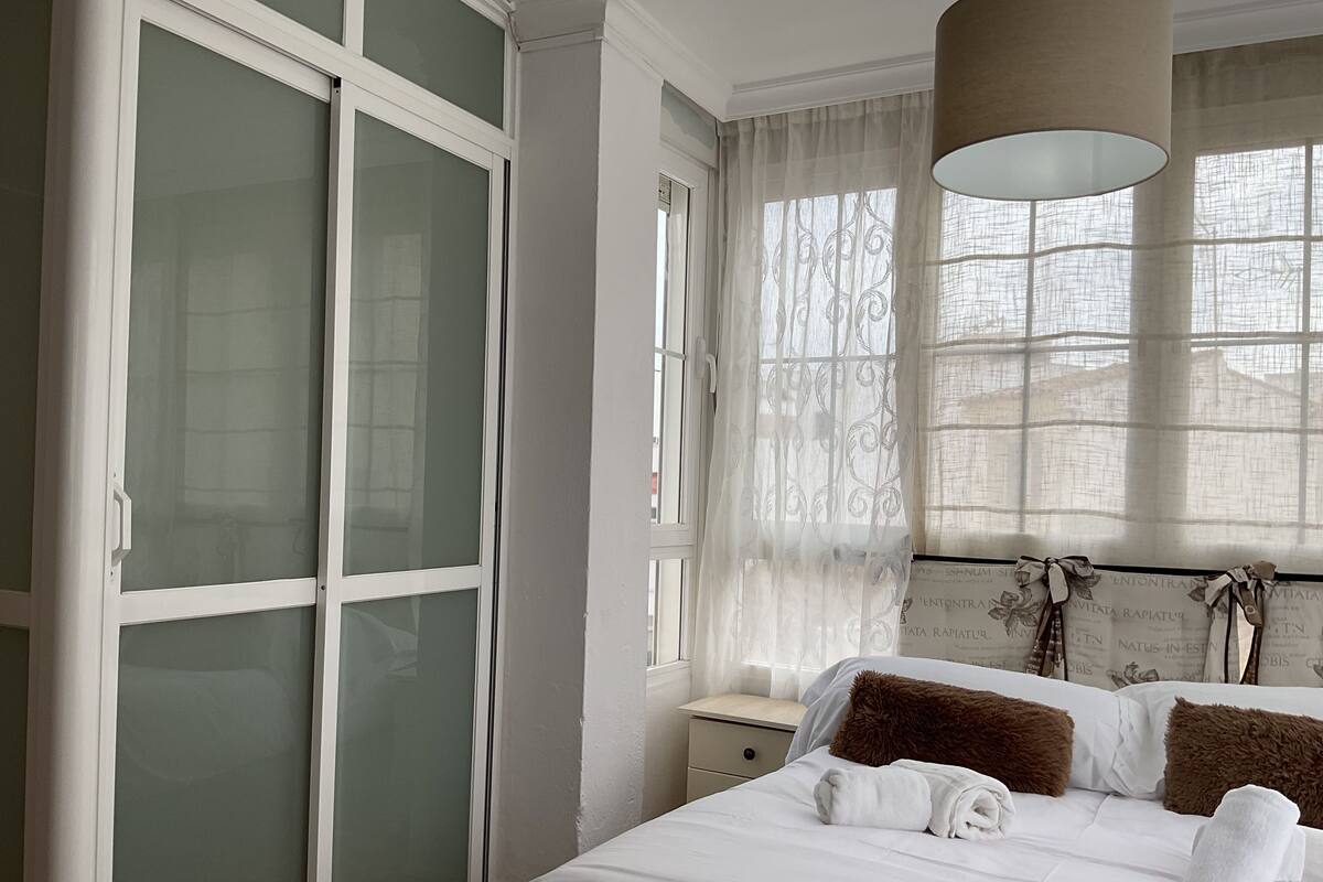 Quarto de hotel com cama, mesa de cabeceira, guarda-roupa e janela com cortinas leves.
