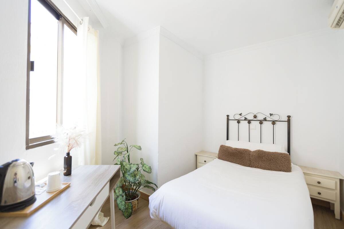 Quarto com cama, mesa de cabeceira, luminária, planta em vaso e janela.