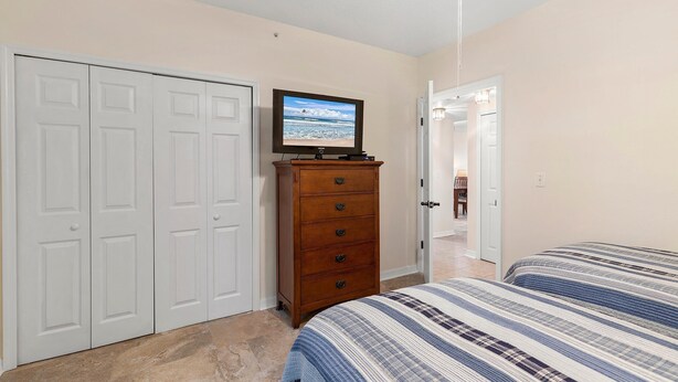 Quarto com cama, cômoda e televisão sobre ela, além de uma porta que dá para outro espaço.