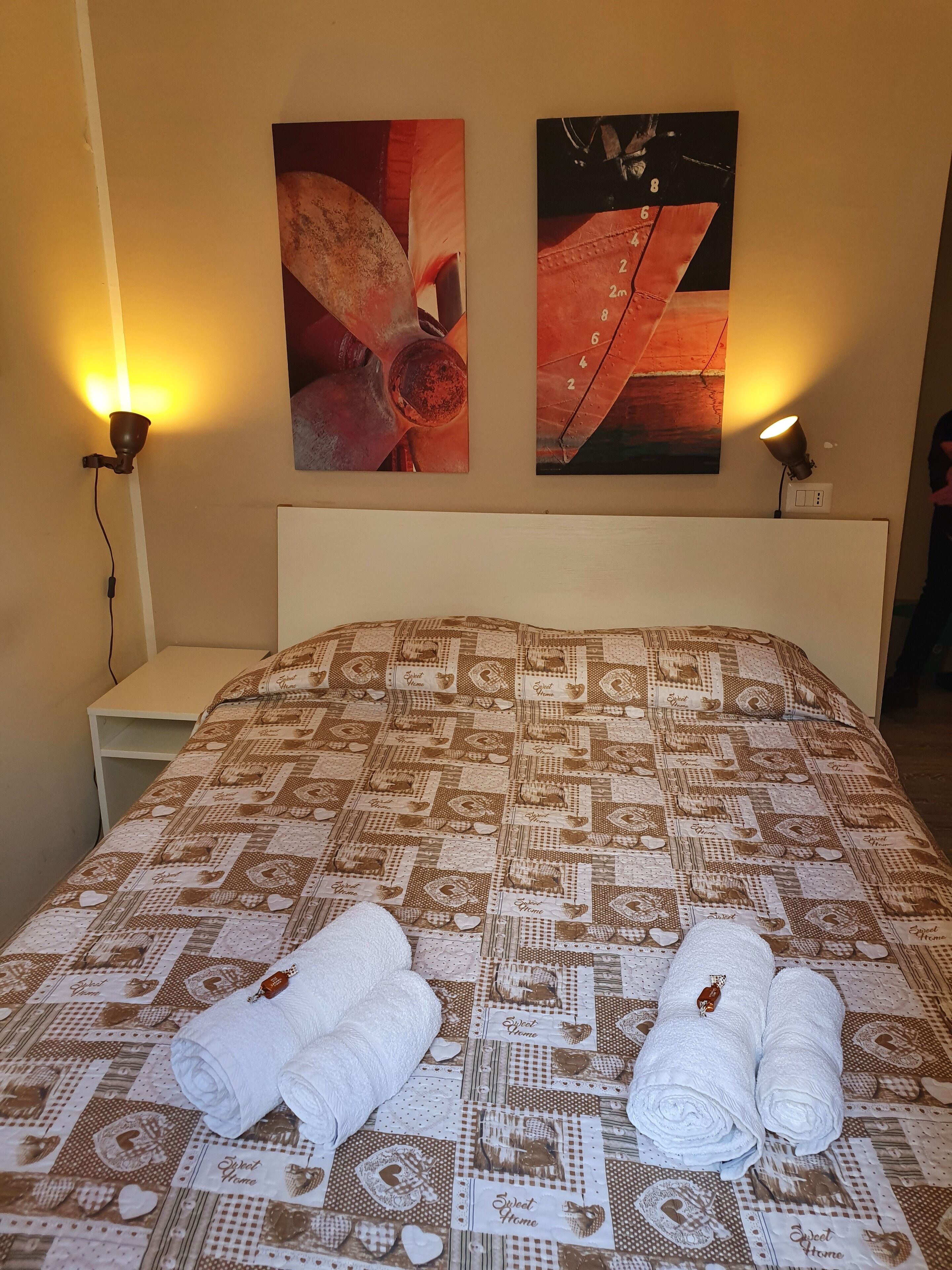 Cama bem arrumada com colcha estampada, dois rolos de toalha e duas luminárias fixadas na parede.
