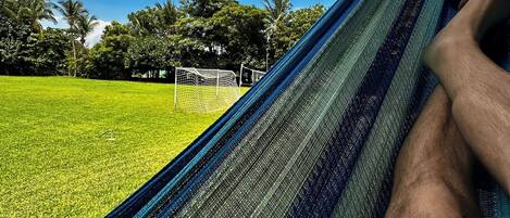 Uma rede com uma pessoa deitada, com vista para um campo gramado e árvores sob um céu parcialmente nublado.