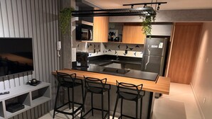 Cozinha moderna com ilha central, banquetas, TV integrada e um espaço de estar com sofá e almofadas.