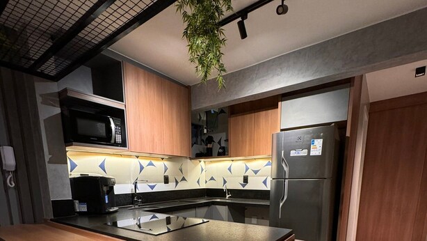 Cozinha moderna com ilha central, armários de madeira, geladeira de aço inoxidável e uma planta pendente.