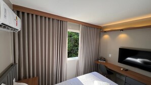 Quarto de hotel com cama, televisão, escrivaninha e janela com cortinas.