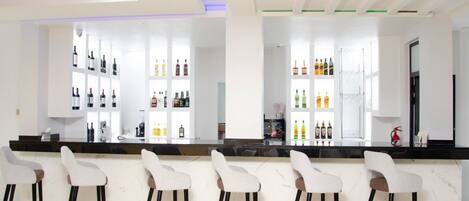 Uma área de bar com banquetas brancas e prateleiras cheias de garrafas diversas.
