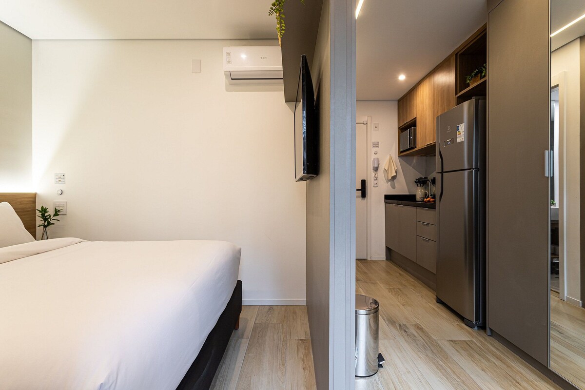 Quarto de hotel moderno com uma cama, uma pequena mesa de cabeceira, uma lixeira, uma TV na parede e uma kitchenette com frigorífico e micro-ondas.