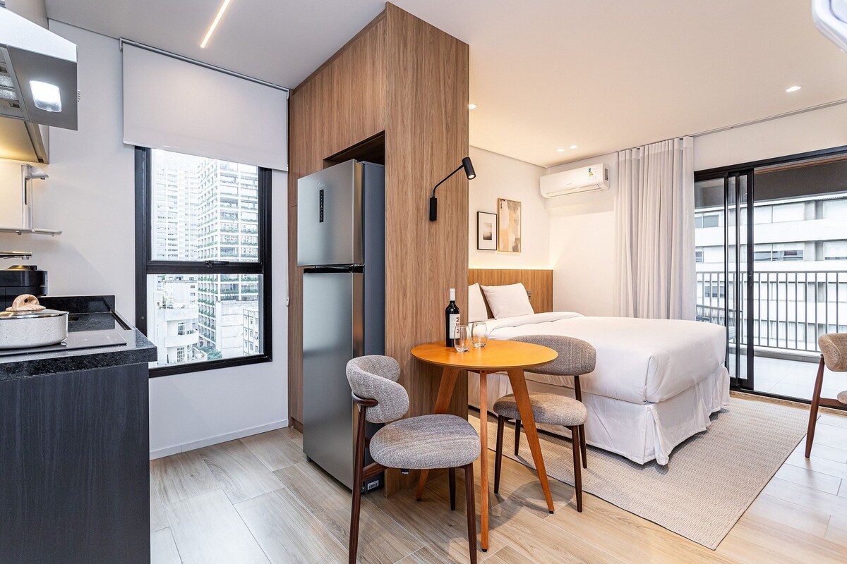 Quarto de hotel moderno com cama, mesa de jantar, duas cadeiras, área de cozinha e uma janela grande com vista para a cidade.