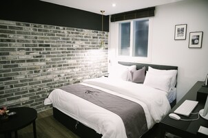 Quarto de hotel moderno com parede de tijolos, uma cama grande com roupa de cama branca, uma escrivaninha com computador e uma janela com persianas.