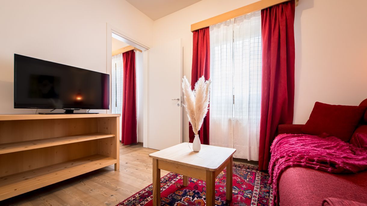 Quarto de hotel moderno com televisão de tela plana em prateleira de madeira, uma mesa pequena com um vaso, colcha vermelha e tapete estampado.