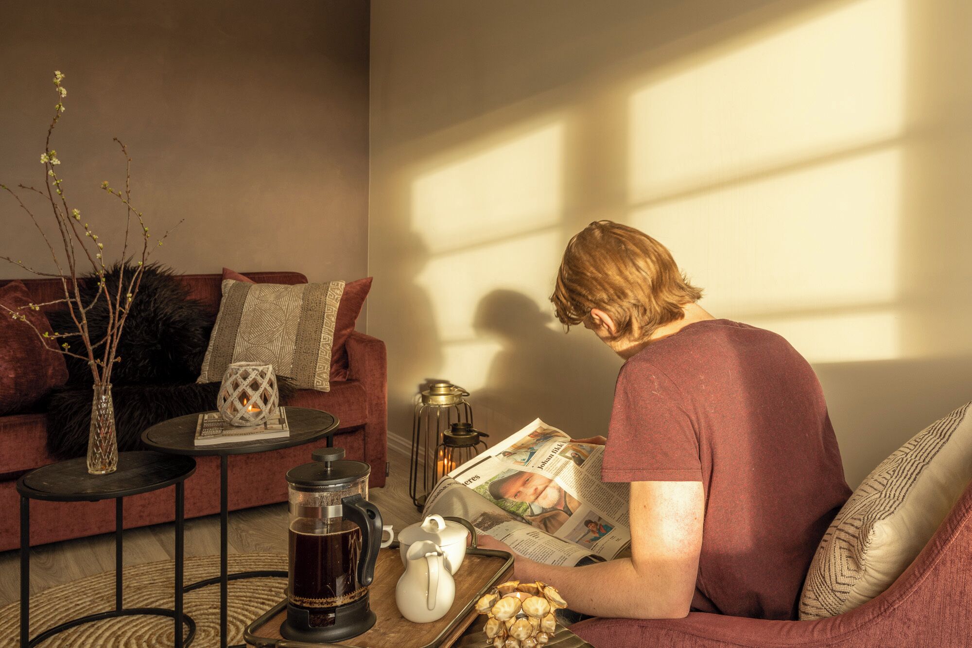 Uma sala de estar aconchegante com um sofá, uma mesa de centro, um vaso com galhos, uma chaleira e um jornal.