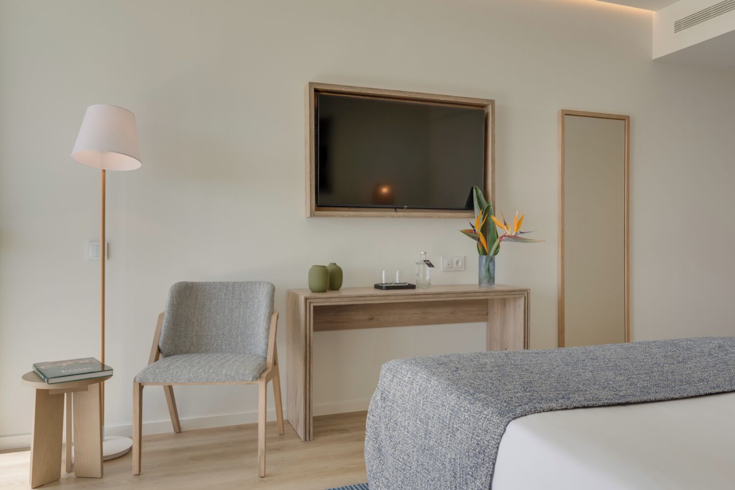 Quarto de hotel com uma cama, uma escrivaninha com um computador, uma cadeira, um abajur e um espelho.