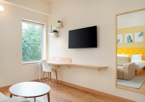 Quarto de hotel moderno com uma grande janela, televisão de tela plana na parede, uma pequena escrivaninha com cadeira e uma mesa redonda.