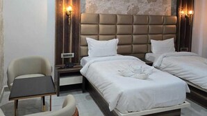 Quarto de hotel com duas camas, uma mesinha, uma cadeira e uma mesinha de cabeceira.