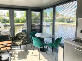 Quarto moderno com área de jantar, cozinha e vista para um corpo de água através de grandes janelas.