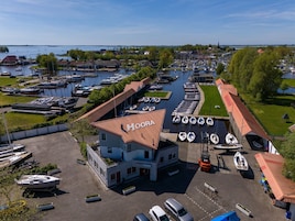 Uma marina com vários barcos atracados, um edifício com a inscrição "Hoora" e uma vista ampla da região circundante, incluindo uma igreja e vegetação.