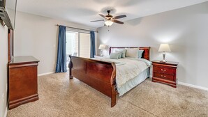 Quarto com cama de madeira, ventilador de teto, mesa de cabeceira com abajur e janela com cortinas.