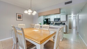 Uma área de refeições com uma mesa de madeira e cadeiras brancas, uma cozinha ao fundo e um corredor que dá para outro quarto.