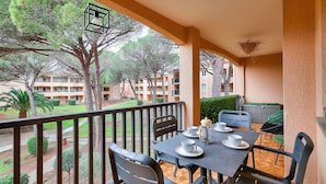 Varanda com mesa posta para dois, cadeiras e vista para um pátio com árvores e edifícios.