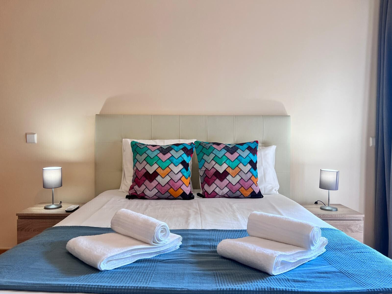 Quarto de hotel com cama, duas almofadas, colcha azul e duas luminárias de cabeceira.
